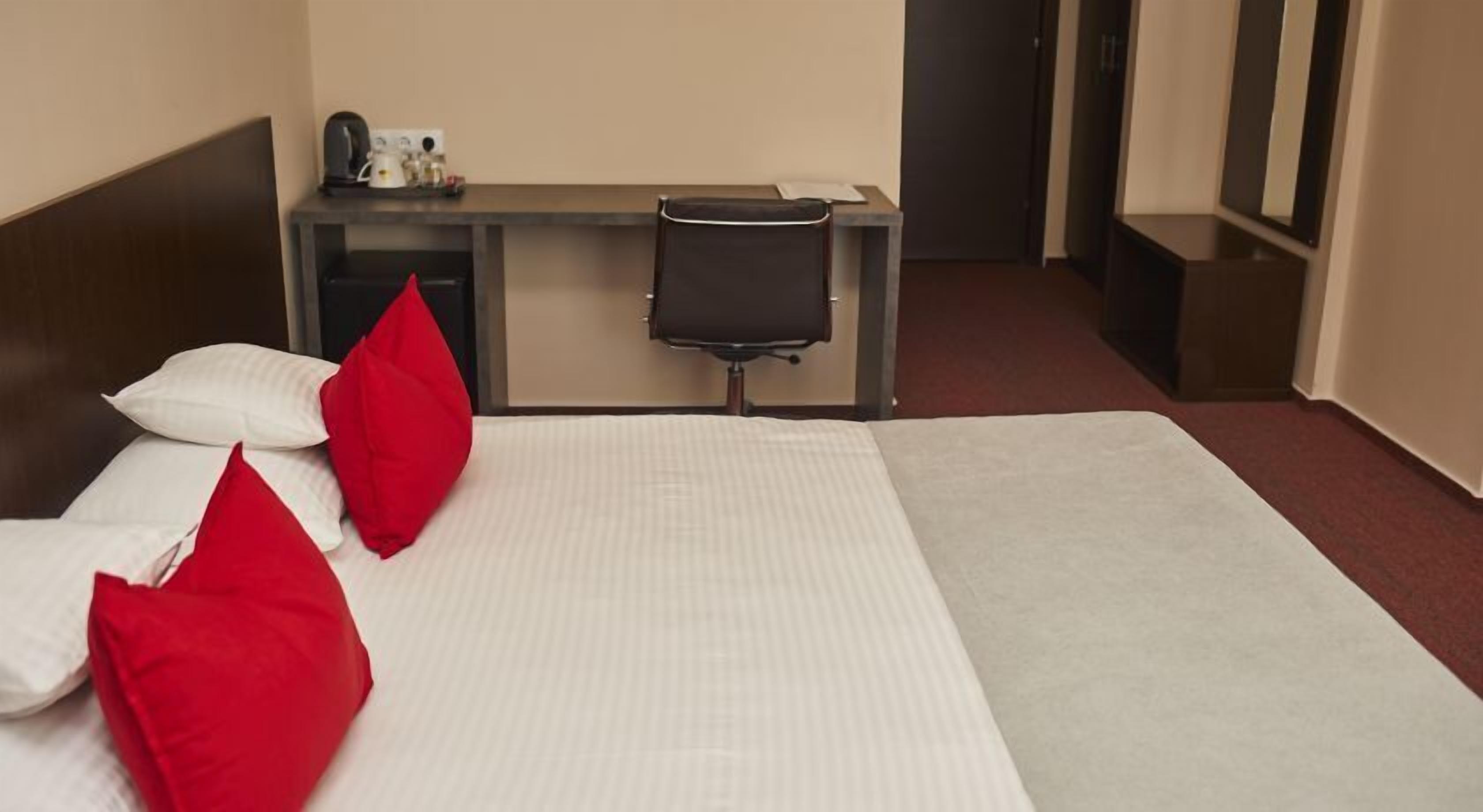 Quarto de hotel com cama, duas almofadas vermelhas, escrivaninha com cadeira e cafeteira.