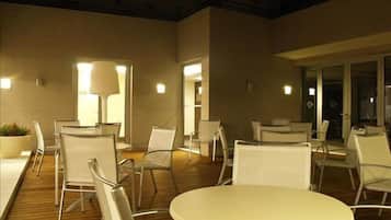 Uma área de refeições ao ar livre com cadeiras brancas e uma mesa redonda, cercada por piso e paredes de madeira.