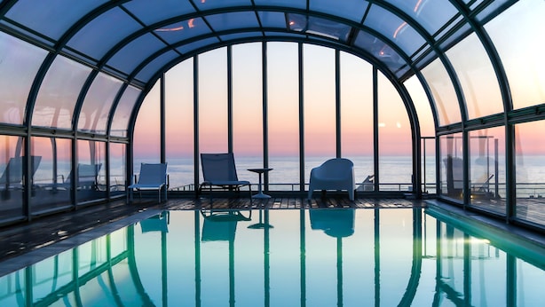 Piscina infinita interior com paredes de vidro transparente, oferecendo vista para o pôr do sol sobre o oceano.
