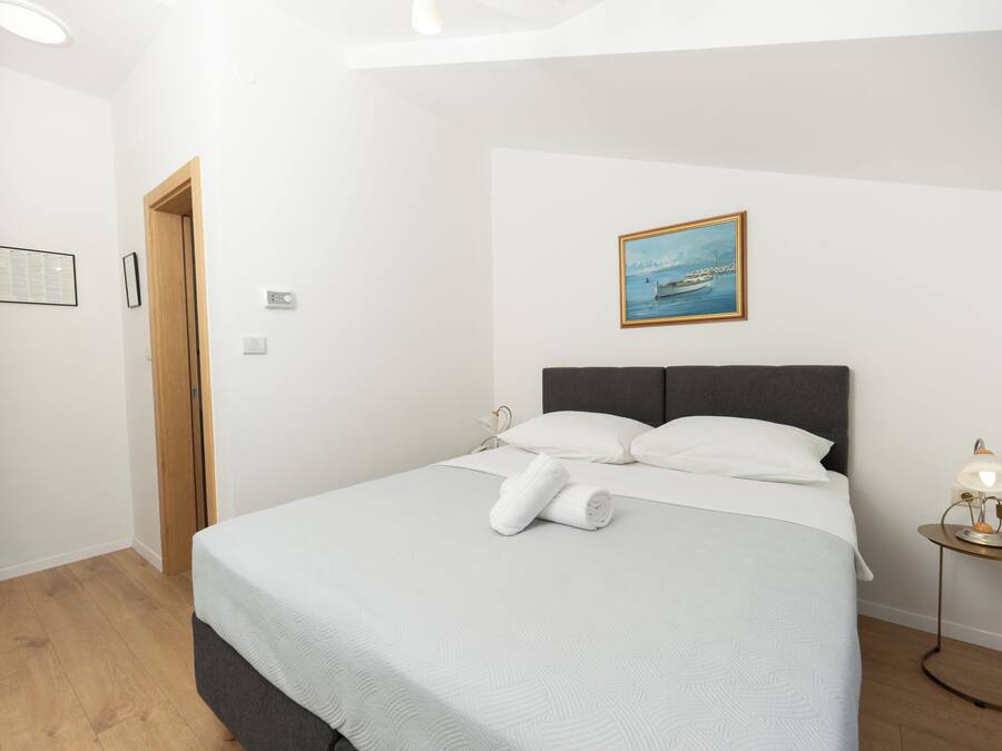 Uma cama bem arrumada com edredom branco e travesseiros, um quadro de um barco na parede, uma pequena mesa de cabeceira com abajur e uma porta de madeira.