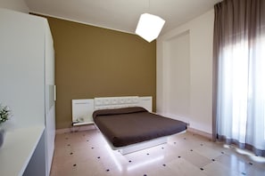 Quarto com cama, mesa de cabeceira, abajur, janela com cortinas e luminária na parede.
