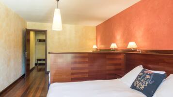 Quarto de hotel com cabeceira de madeira, cama com lençóis brancos, duas luminárias de apoio e uma porta que dá para um corredor.