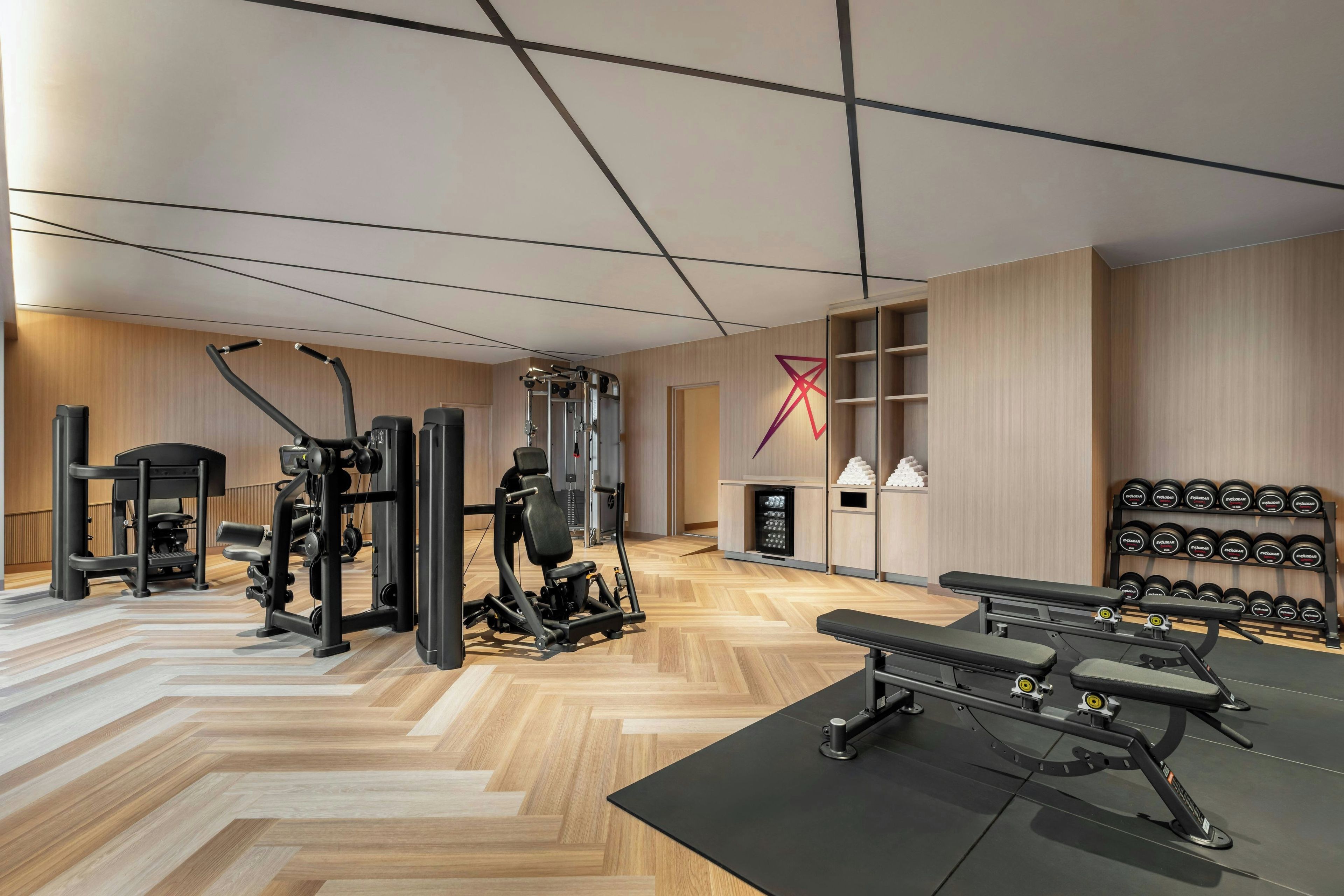 Academia com diversos equipamentos de exercícios, piso em padrão herringbone e suporte de halteres fixado na parede.