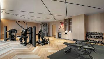 Academia com diversos equipamentos de exercícios, piso em padrão herringbone e suporte de halteres fixado na parede.
