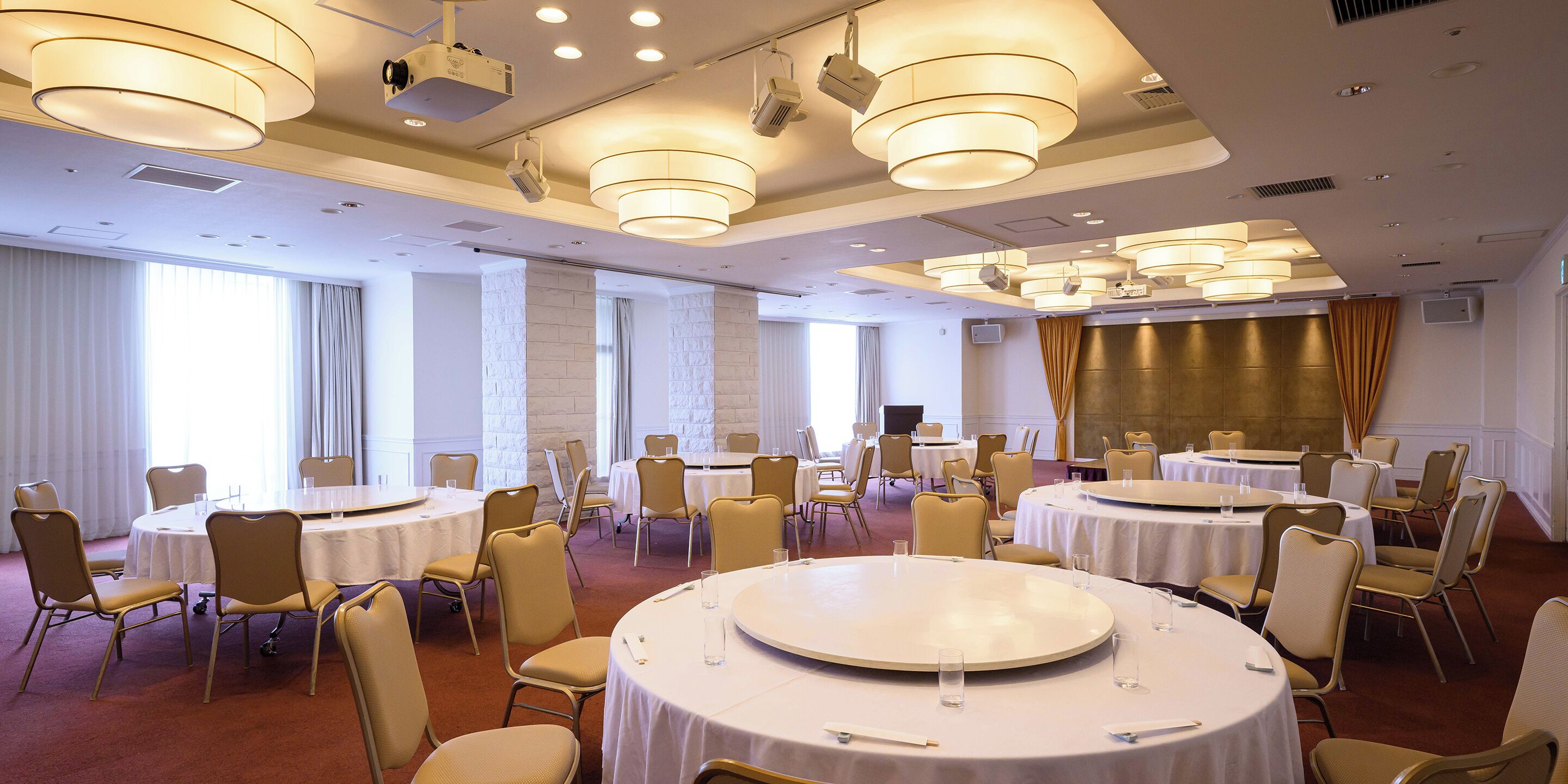 Sala de conferências com várias mesas redondas, cadeiras bege e luminárias modernas.