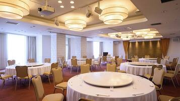 Sala de conferências com várias mesas redondas, cadeiras bege e luminárias modernas.