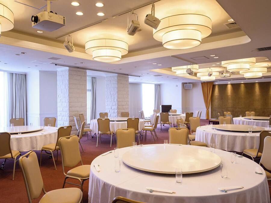 Sala de conferências com várias mesas redondas, cadeiras bege e luminárias modernas.