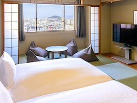 Quarto de hotel com cama, uma pequena mesa, duas cadeiras, televisão e vista para o horizonte urbano através de grandes janelas.
