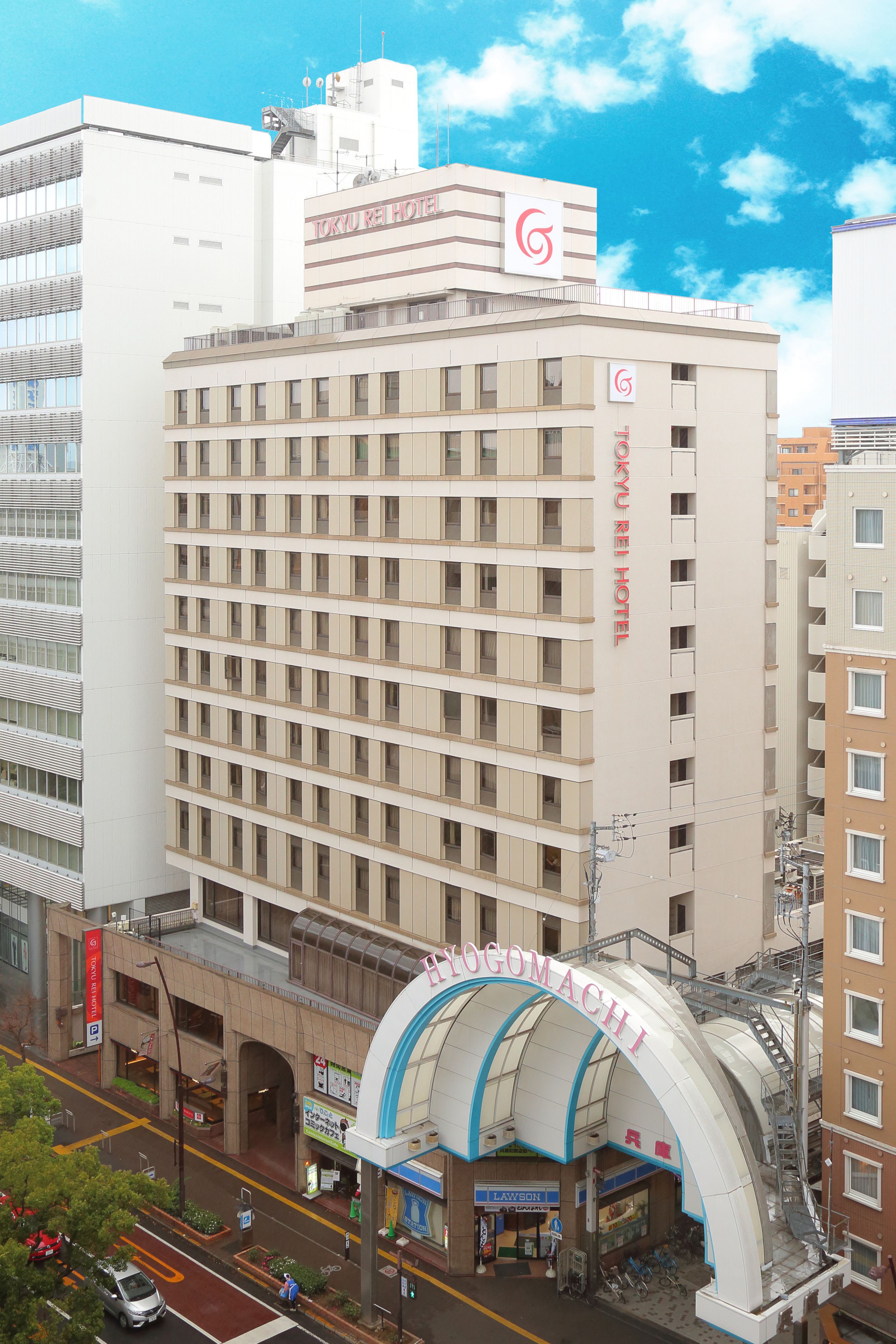 Um hotel de vários andares com uma placa de destaque "TOKYU REI HOTEL", uma grande entrada em arco e uma área de estacionamento visível abaixo.