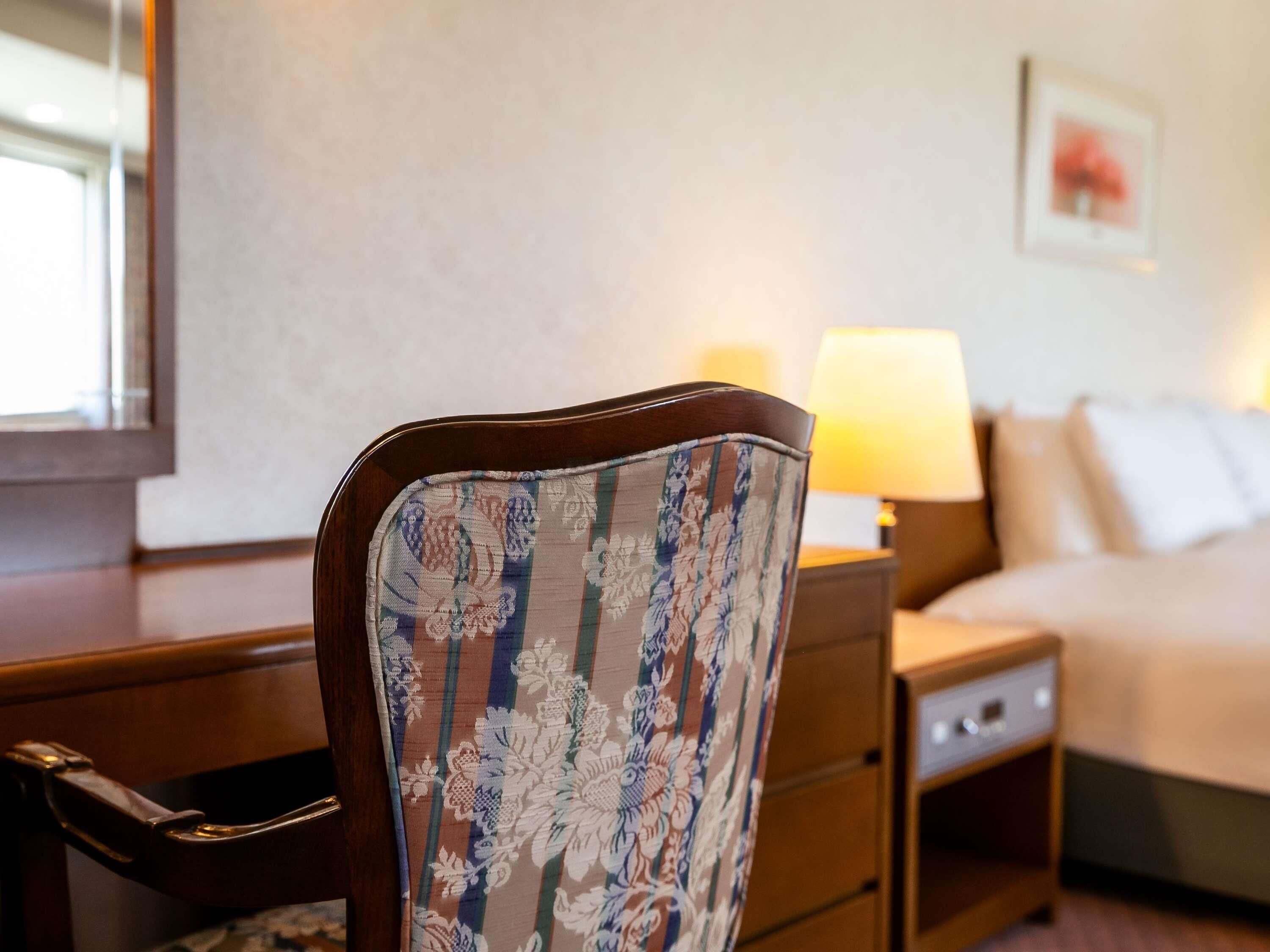 Quarto de hotel com cama, mesa de cabeceira, luminária e poltrona estampada com flores.