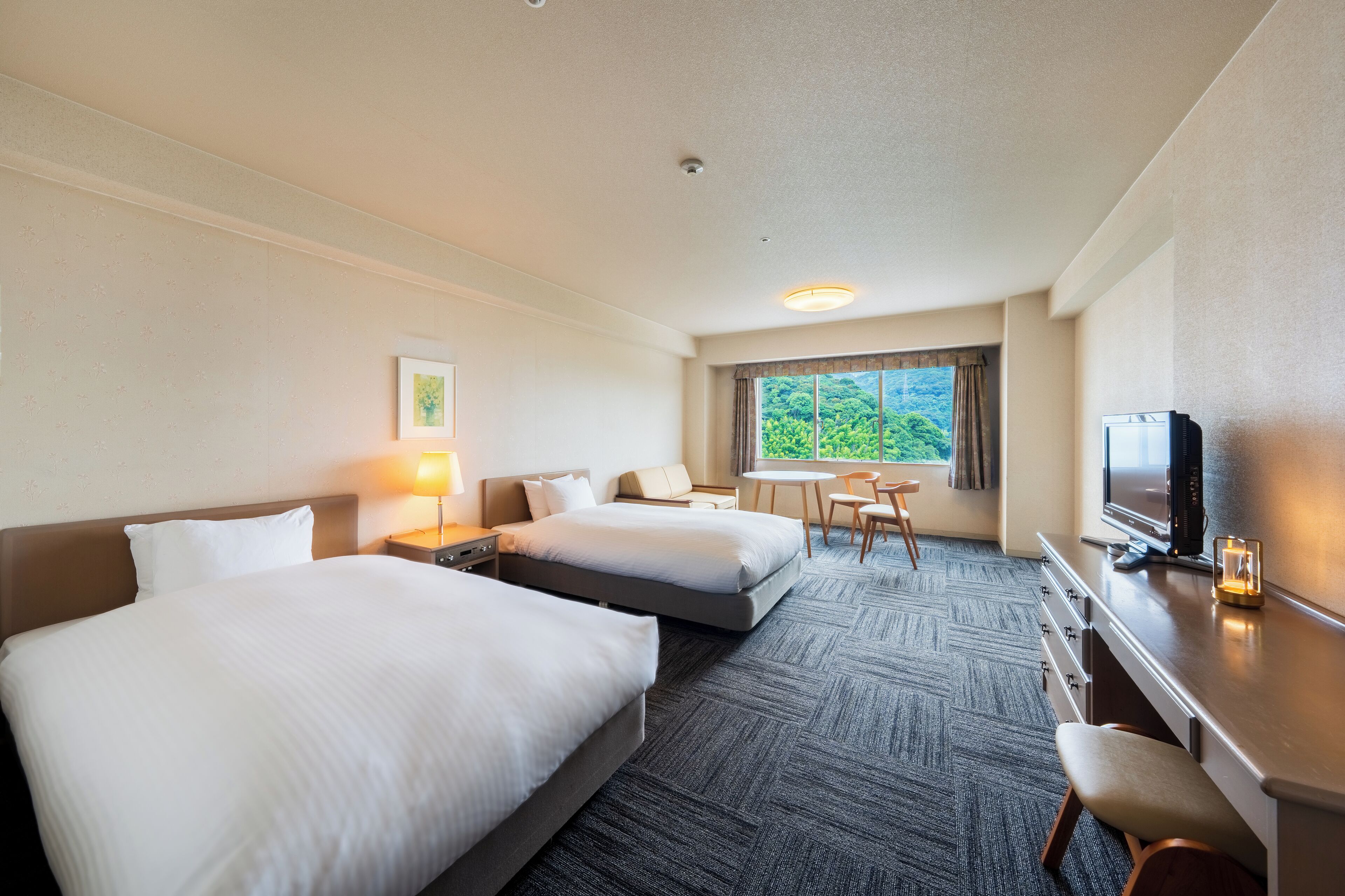 Quarto de hotel com duas camas, uma mesa de trabalho com televisão, uma cadeira e uma janela com vista para vegetação.