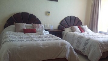 Quarto de hotel com duas camas, cada uma com um design de cabeceira diferente, uma mesa de cabeceira com abajur e uma janela com cortinas.