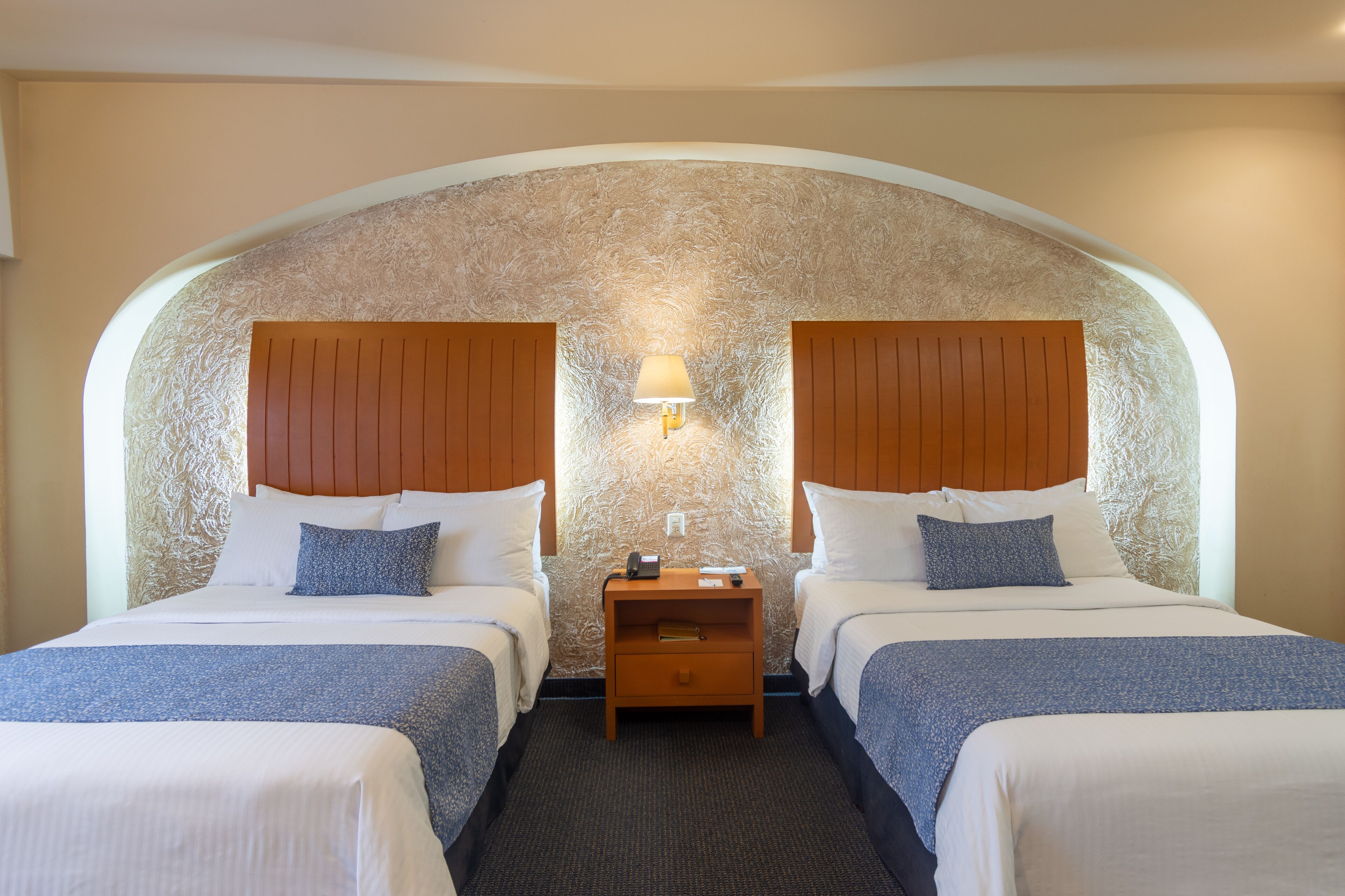 Quarto de hotel com duas camas, uma mesinha de cabeceira e uma parede com padrão texturizado.