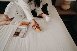 Uma pessoa sentada na cama com um livro, um prato com um croissant e uma xícara.