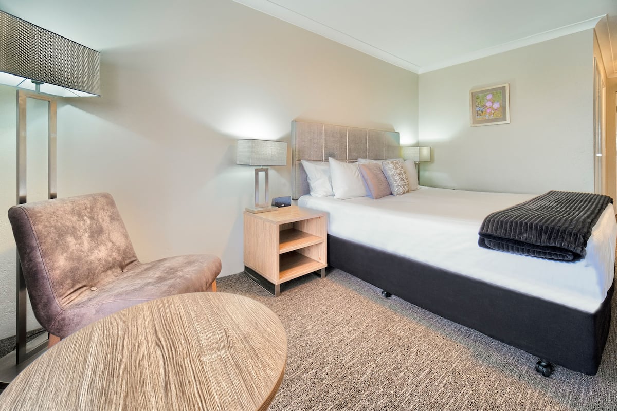 Quarto de hotel com cama, uma cadeira, mesa pequena, mesa de cabeceira e luminária.