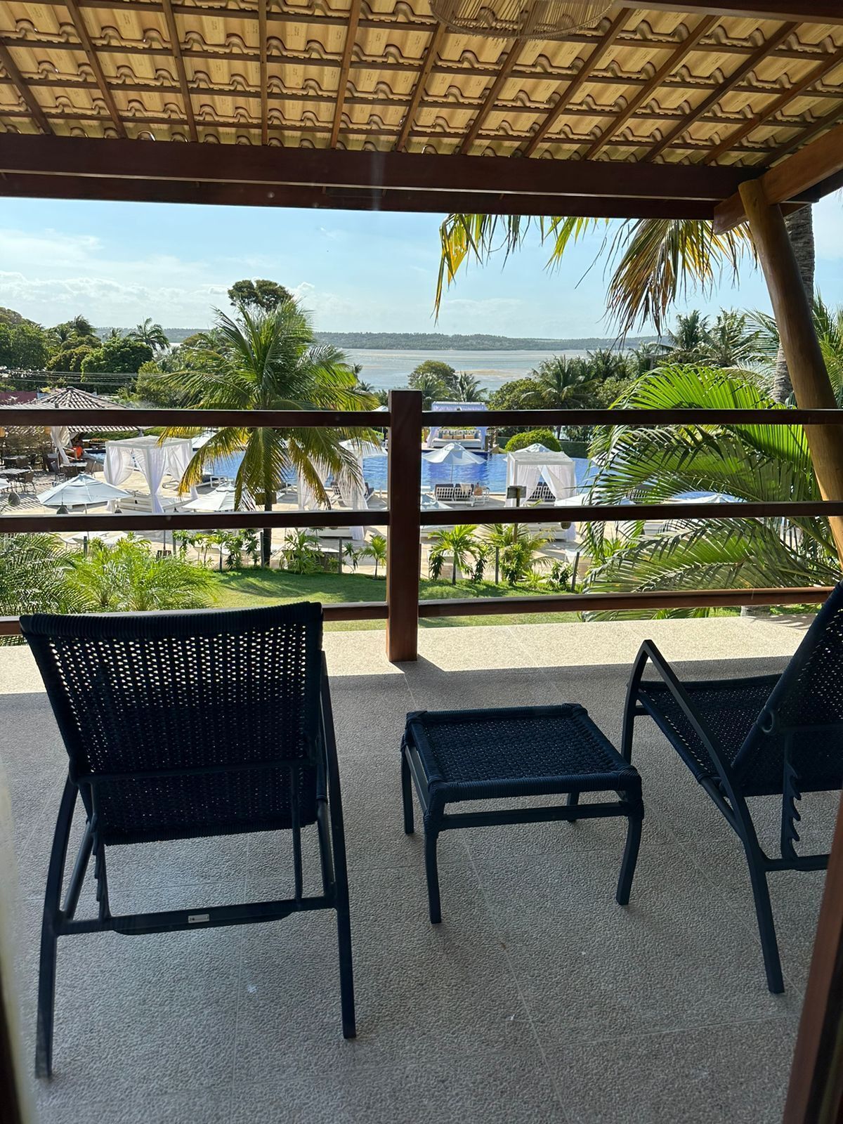 Uma área de estar coberta ao ar livre, com duas cadeiras e uma pequena mesa, com vista para uma praia com guarda-sóis e palmeiras.