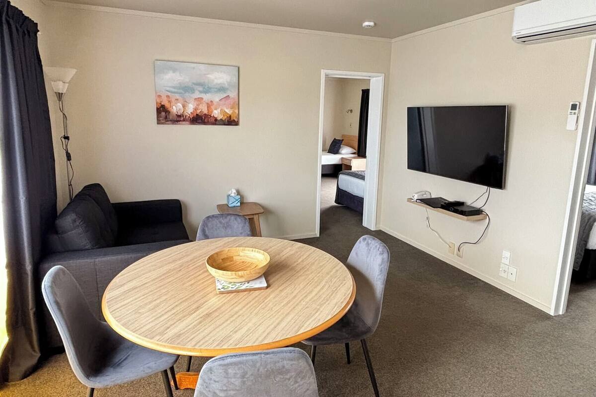 Quarto de hotel com uma mesa redonda de madeira, cadeiras estofadas cinzentas, uma televisão de tela plana na parede, um quadro na parede e uma cama ao fundo.