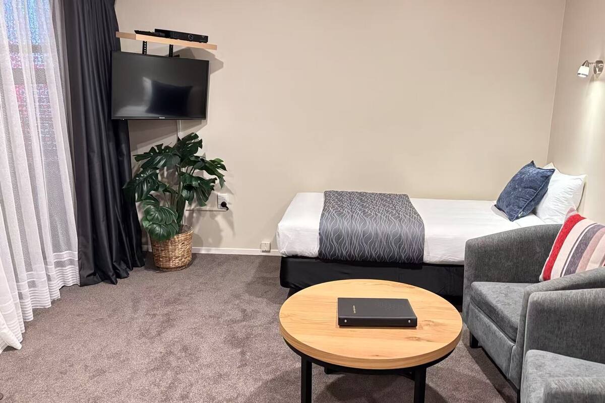 Quarto de hotel moderno com cama, sofá cinza, mesa de centro redonda de madeira, televisão fixada na parede e planta em vaso.