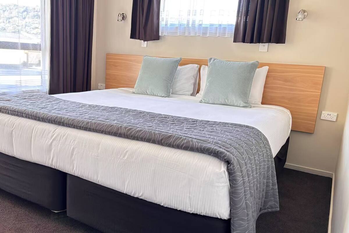 Uma cama bem arrumada com colcha cinza e branca estampada, duas almofadas verde-claras e cabeceira de madeira.