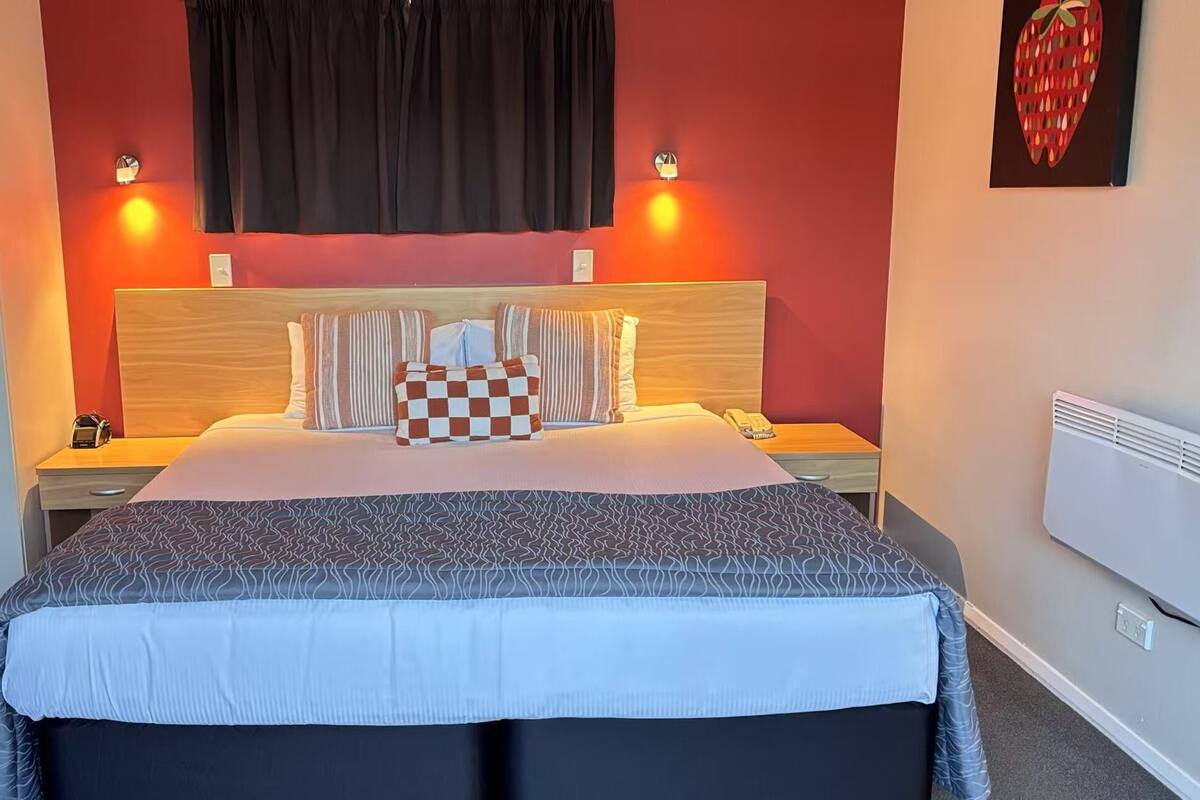 Quarto de hotel com uma cama grande, duas mesas de cabeceira, um criado-mudo, um relógio de parede, um aquecedor e uma parede vermelha com um objeto decorativo.