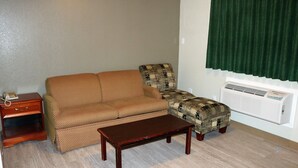 Uma sala de estar com um sofá marrom, uma poltrona estampada, uma mesa de centro de madeira, um criado-mudo de madeira pequeno e um ar-condicionado fixado na parede.
