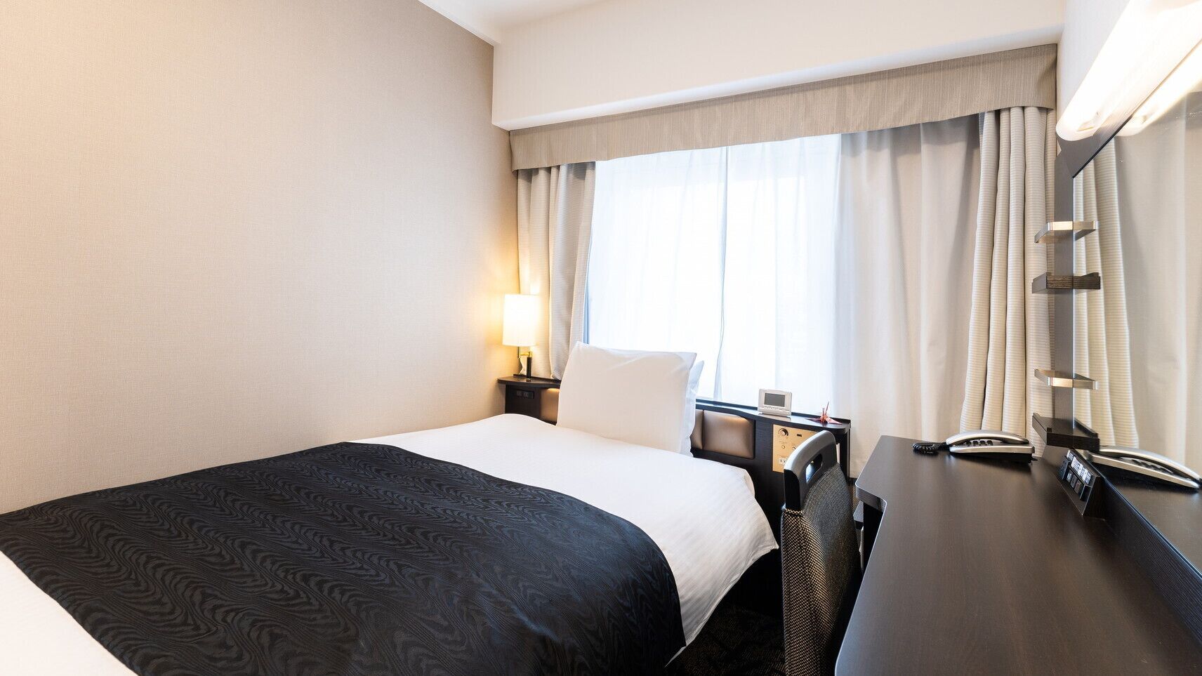 Quarto de hotel com cama, mesa de cabeceira, luminária e escrivaninha com telefone.