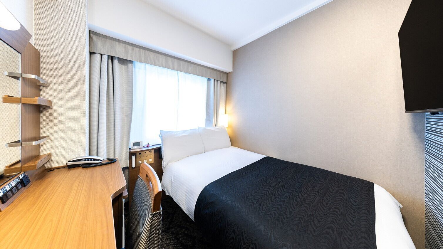 Quarto de hotel com cama, escrivaninha com telefone, cadeira, mesa de cabeceira e janela com cortinas.