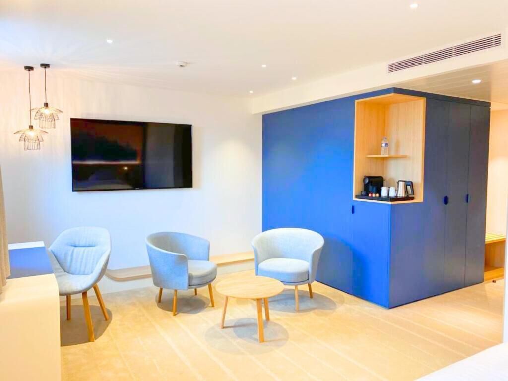 Quarto de hotel moderno com televisão de tela plana, três cadeiras, uma mesa redonda pequena e um armário azul com prateleira.