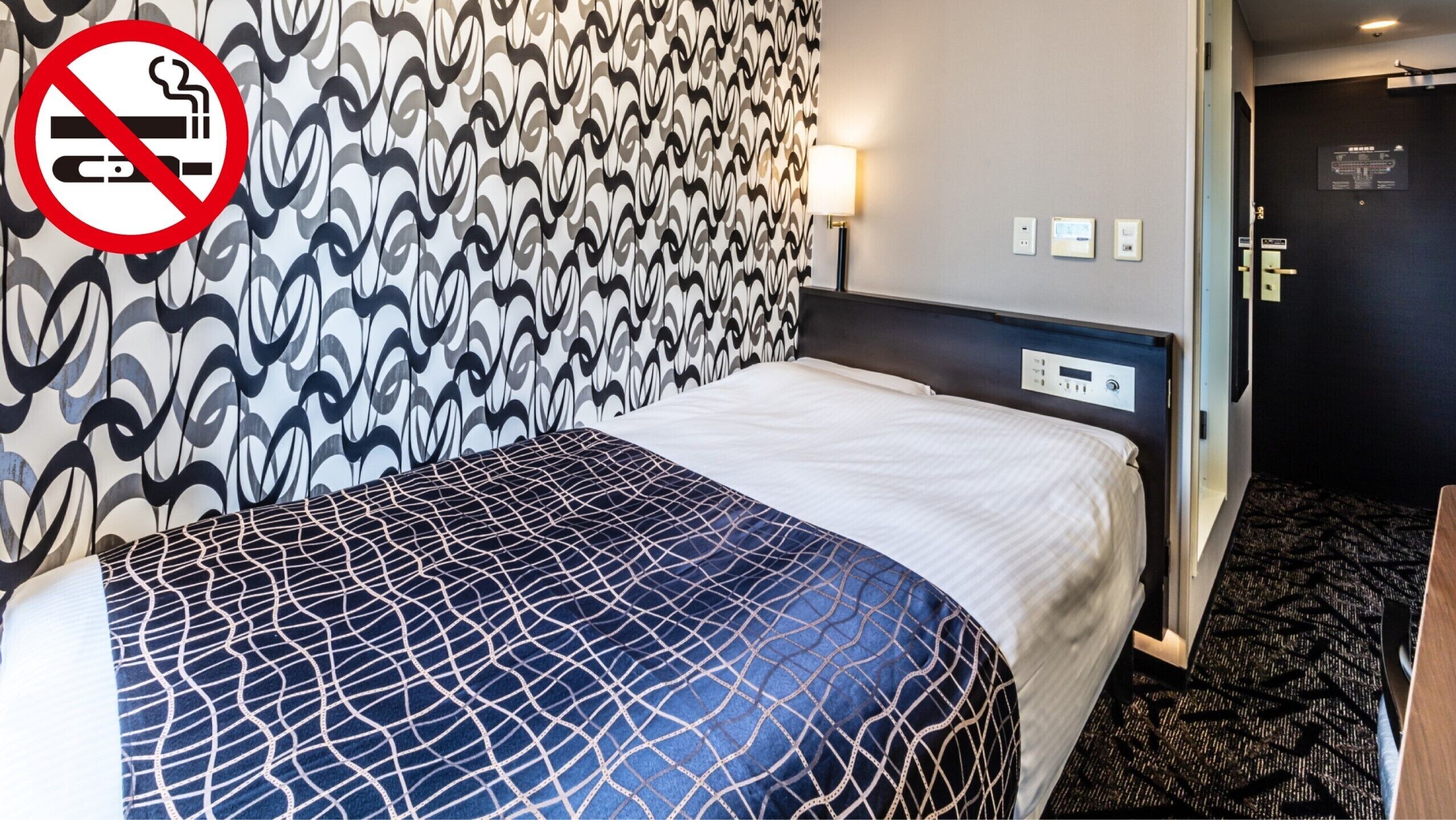 Quarto de hotel com parede estampada, cama com colcha azul e branca, televisão fixada na parede e placa indicando proibido fumar.