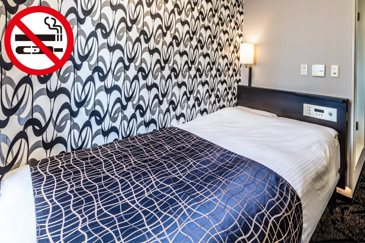 Quarto de hotel com parede estampada, cama com colcha azul e branca, televisão fixada na parede e placa indicando proibido fumar.