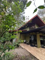 Uma casa moderna com telhado vermelho, varanda coberta e área de jardim com árvores e plantas.