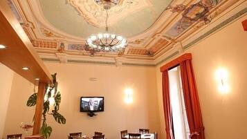Uma sala de jantar com pé-alto, detalhes ornamentais no teto e um candelabro. Mesas montadas com cadeiras, e uma televisão fixada na parede.