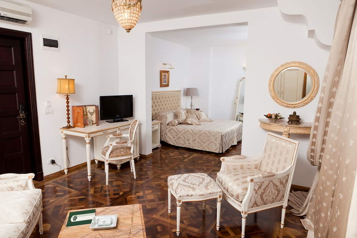Quarto de hotel com cama, escrivaninha com computador, cadeira, mesa pequena, espelho e lustre.