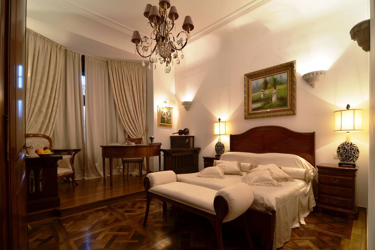 Quarto de hotel com uma cama grande, uma escrivaninha com cadeira, um lustre e um quadro na parede.