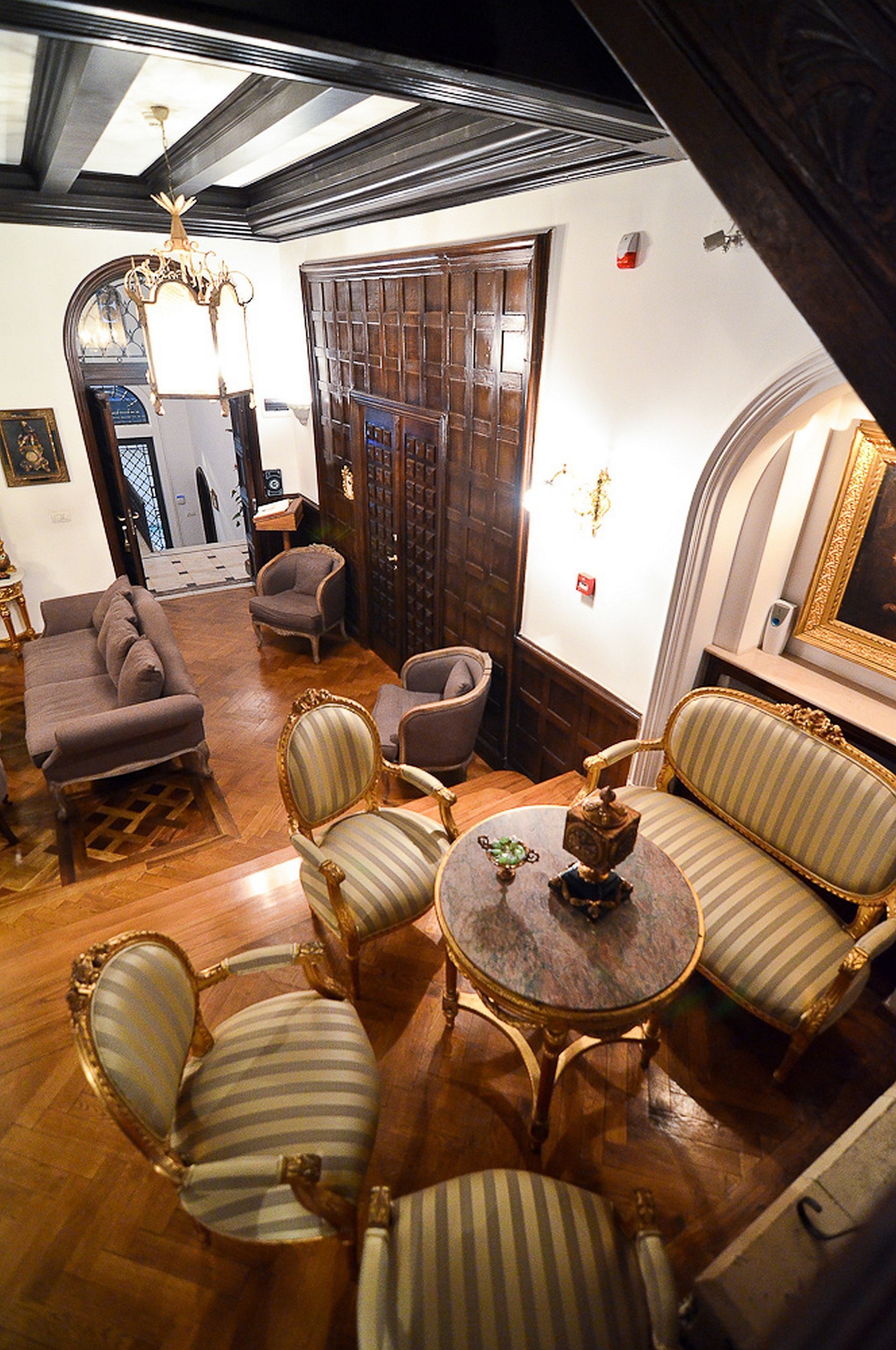 Quarto com piso de madeira, um sofá, cadeiras listradas, uma mesa com um objeto decorativo e uma grande porta de madeira.