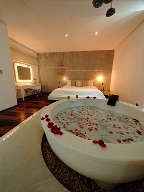Quarto com uma banheira grande cheia de água e pétalas de rosa, toucador com espelho e iluminação, e cama com almofadas.