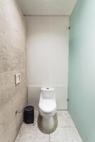 Casa de banho pequena e compacta, com sanita branca, um lixo preto e chão em mosaico claro.