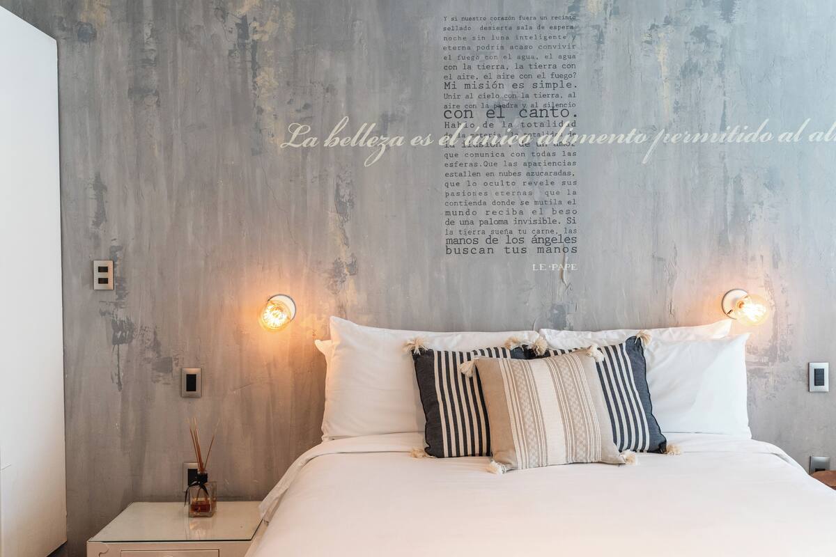 Quarto com cama, duas luminárias na parede, mesa de cabeceira e parede com uma frase.