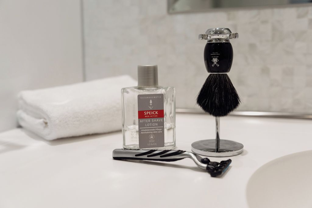 Casa de banho com lavatório branco, frasco de loção pós-barba Speick Men Active, barbeador de segurança preto e toalha branca.