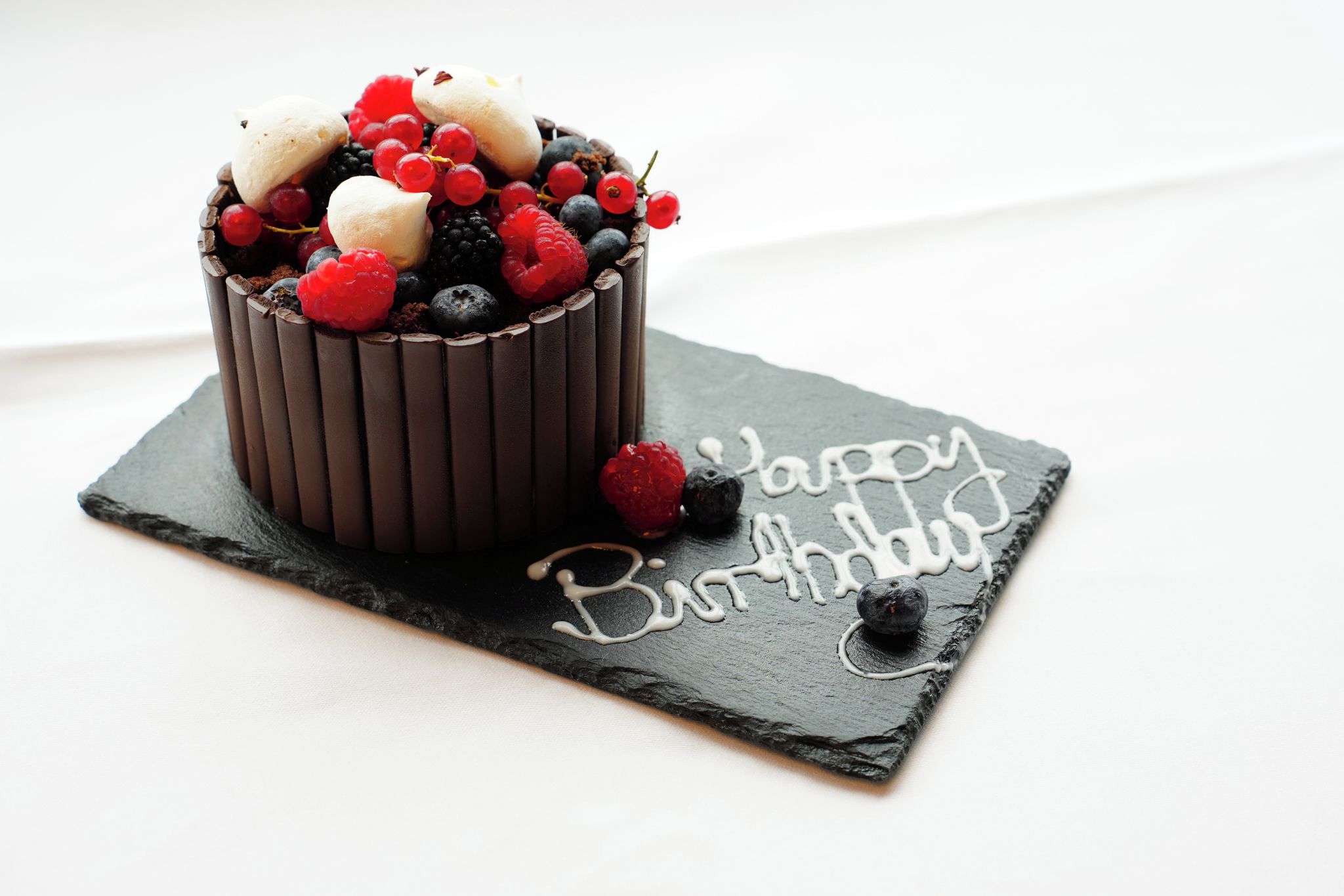 Bolo de chocolate com frutos vermelhos sobre uma tábua de ardósia com a mensagem 'Happy Birthday'.