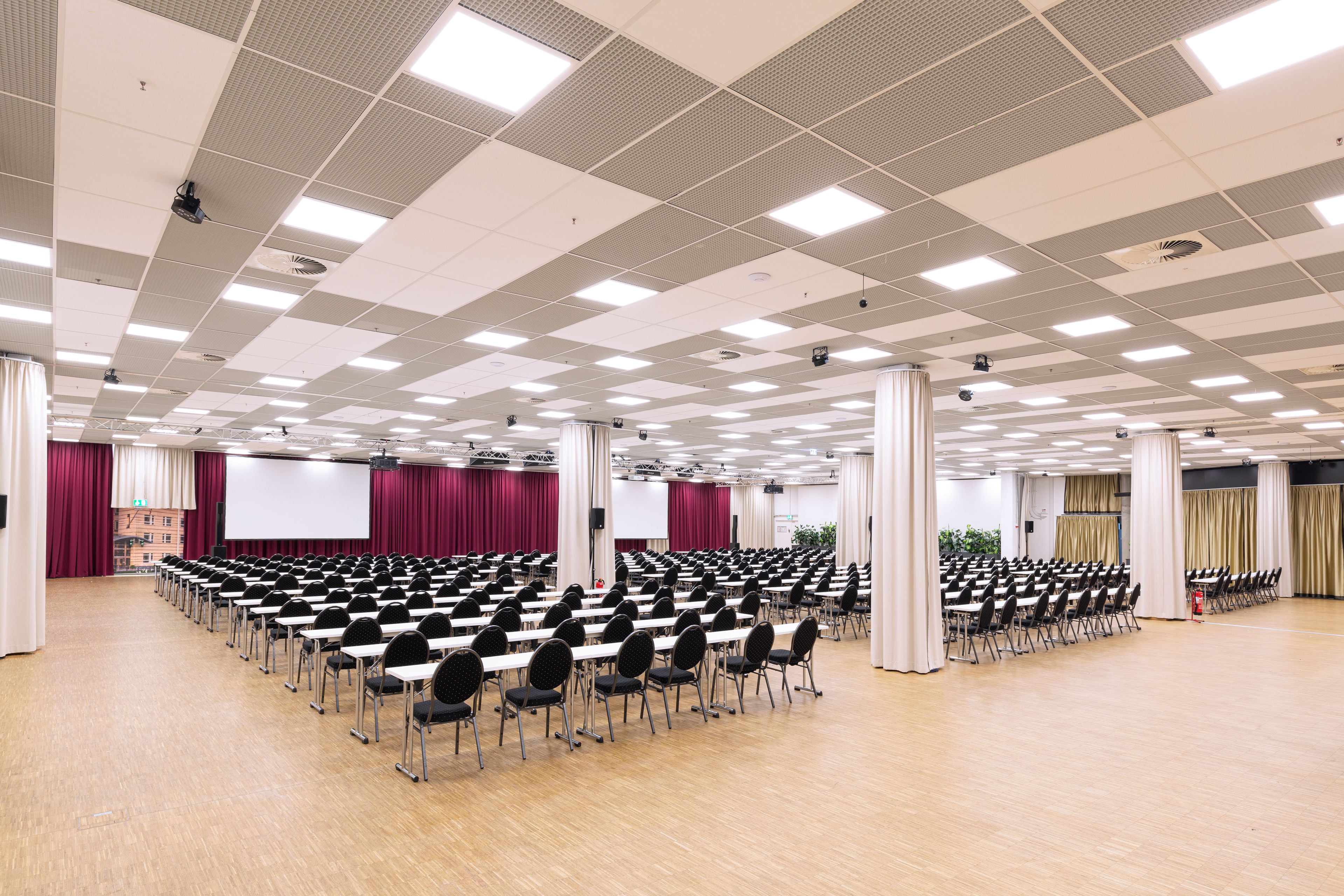Uma grande sala de conferências com fileiras de cadeiras, palco com tela e alto teto com luminárias.