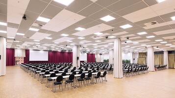 Uma grande sala de conferências com fileiras de cadeiras, palco com tela e alto teto com luminárias.