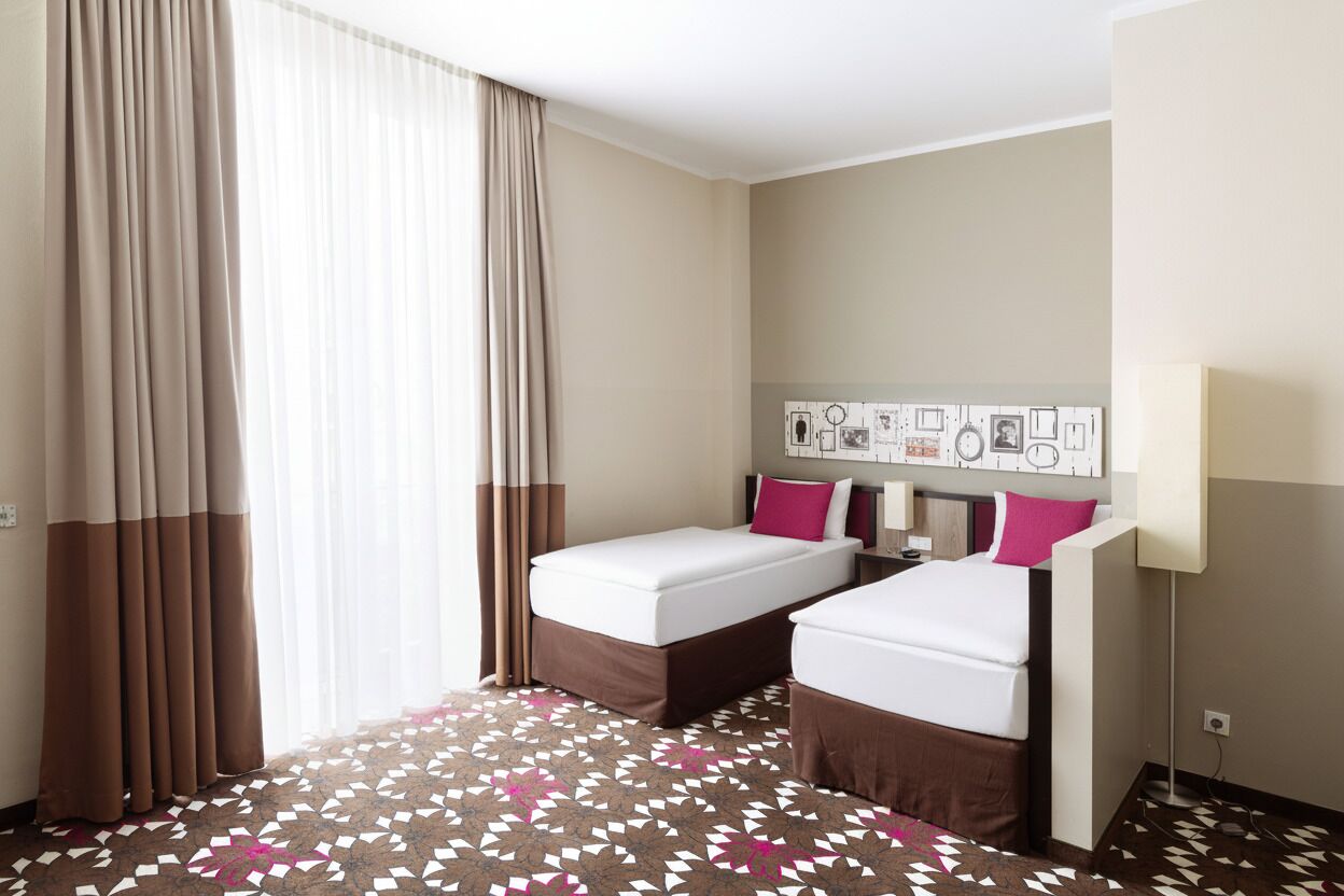 Quarto de hotel com duas camas, uma janela grande com cortinas, um chão estampado e uma parede com quadros.