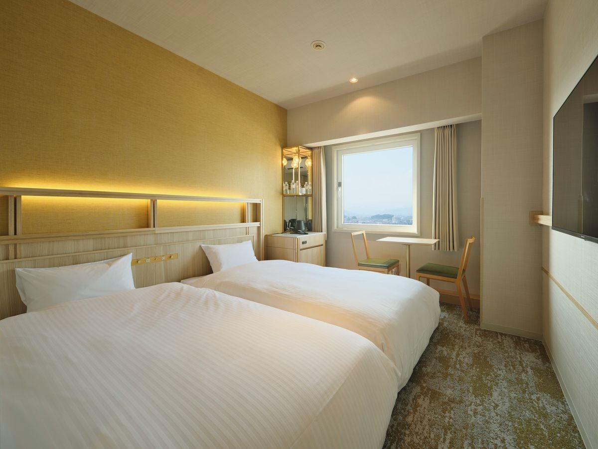 Quarto de hotel com duas camas, uma escrivaninha, uma cadeira, uma televisão e uma janela com cortinas.