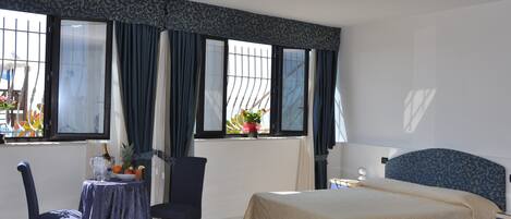 Quarto de hotel com uma cama, uma mesa com uma cesta de frutas, uma cadeira e uma janela com cortinas.