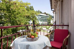 Uma varanda com mesa posta, com frutas e copos de vinho, uma cadeira de vime e vista para um edifício e árvores.