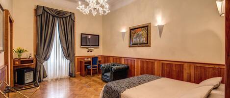 Quarto de hotel com uma cama grande, uma escrivaninha com cadeira, televisão e um candelabro.