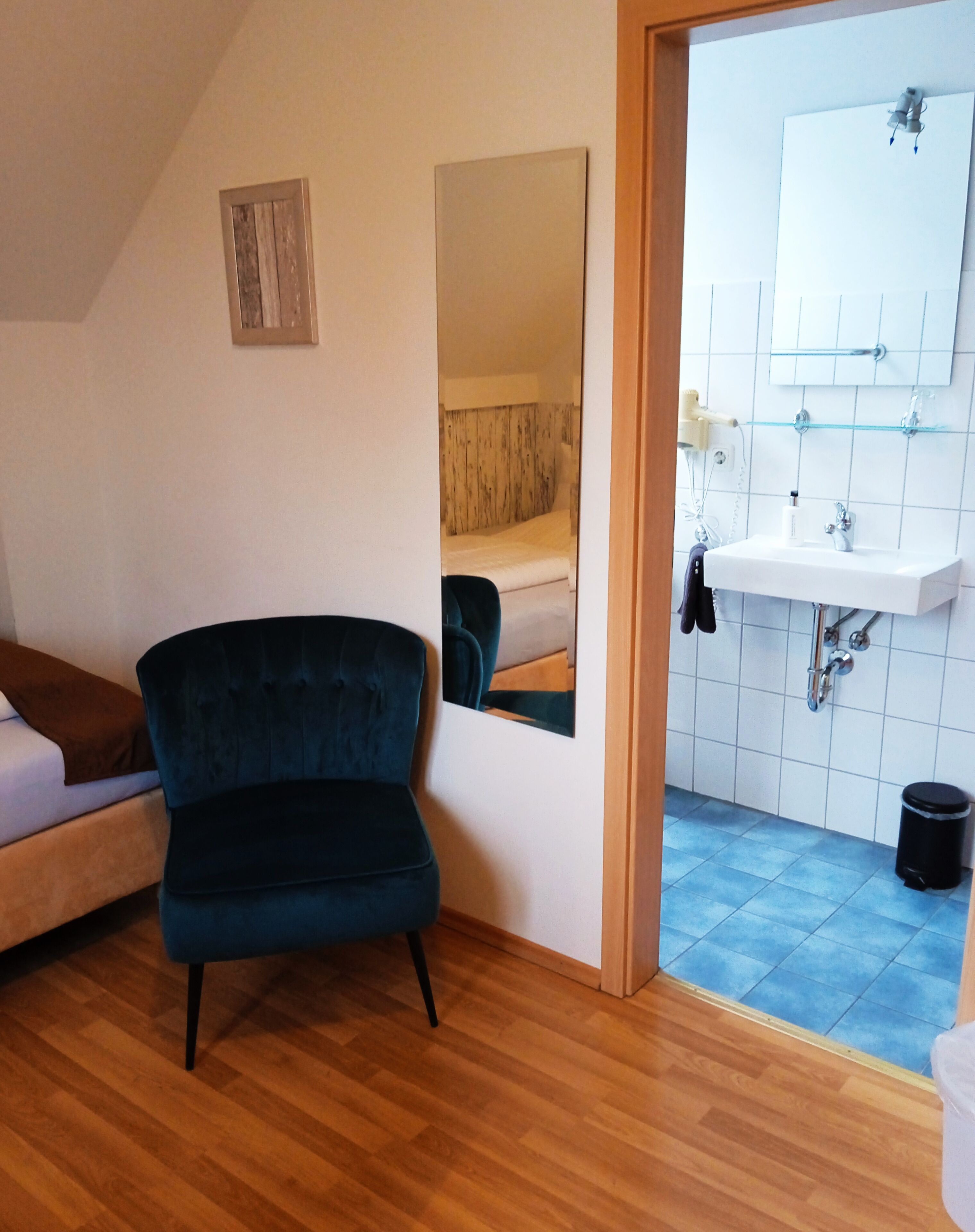 Um quarto com piso de madeira, uma cadeira azul escura, uma cama com cabeceira de madeira, um espelho, uma pia branca e uma lixeira azul.