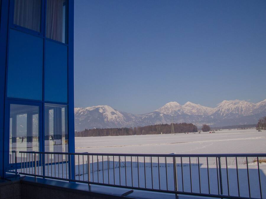 Vista de uma varanda com grade de metal, com vista para uma paisagem coberta de neve e montanhas.