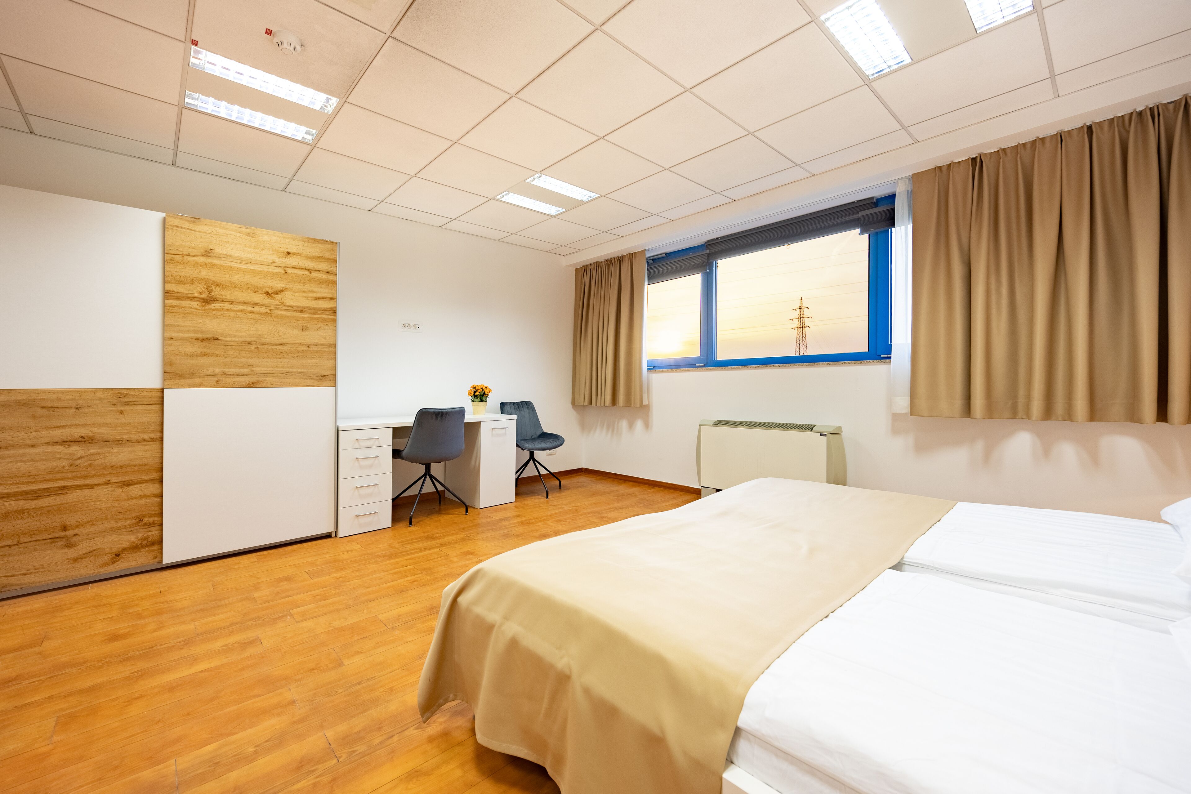 Quarto de hotel com parede de destaque em madeira, uma mesa com cadeira, cama com colcha bege e janela com cortinas.